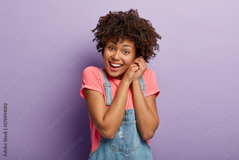 Smiling friendly dark skinned woman keeps hands gently together near ...