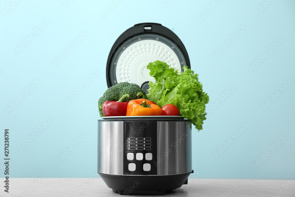 Modern multi cooker with vegetables on table against color background ...