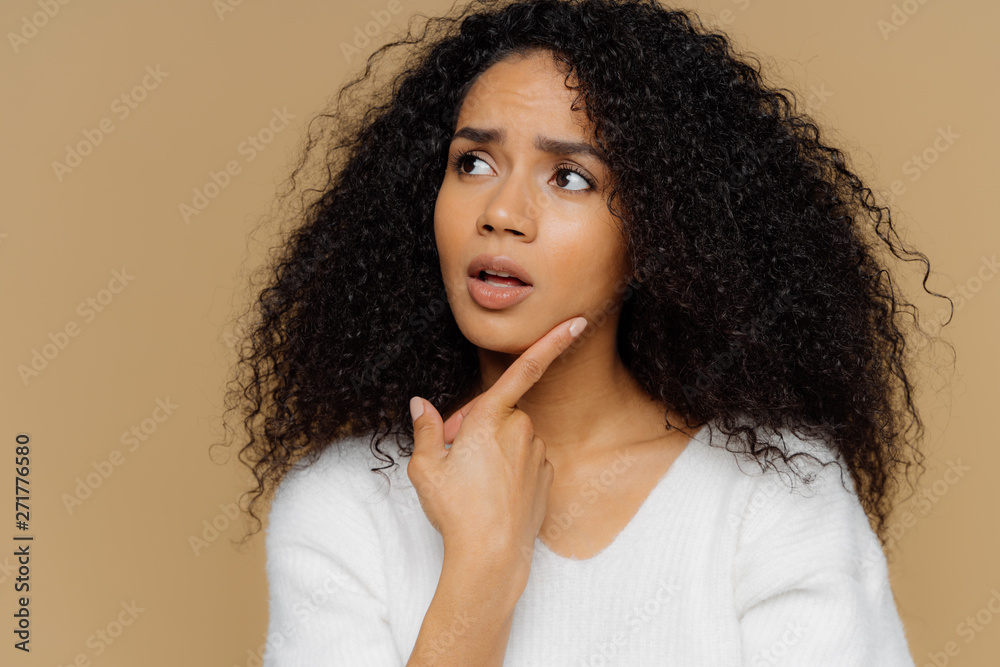 Puzzled female model keeps fore finger on chin, looks thoughtfully ...