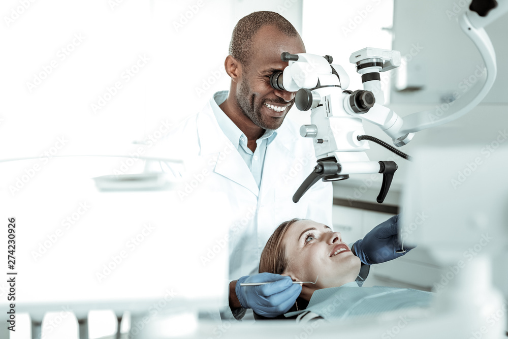 Dark-skinned doctor wearing uniform and looking in special microscope ...