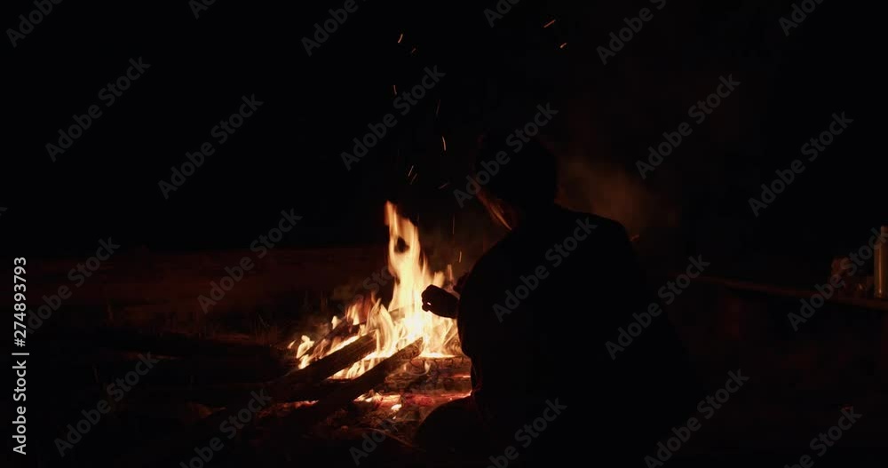 Female traveler sitting alone by fire in evening warming hands ...