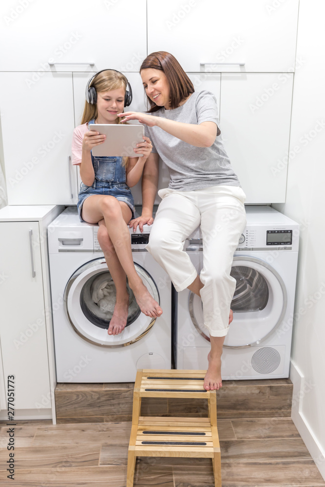 Young mother and her daughter are playing with tablet in laundry room ...