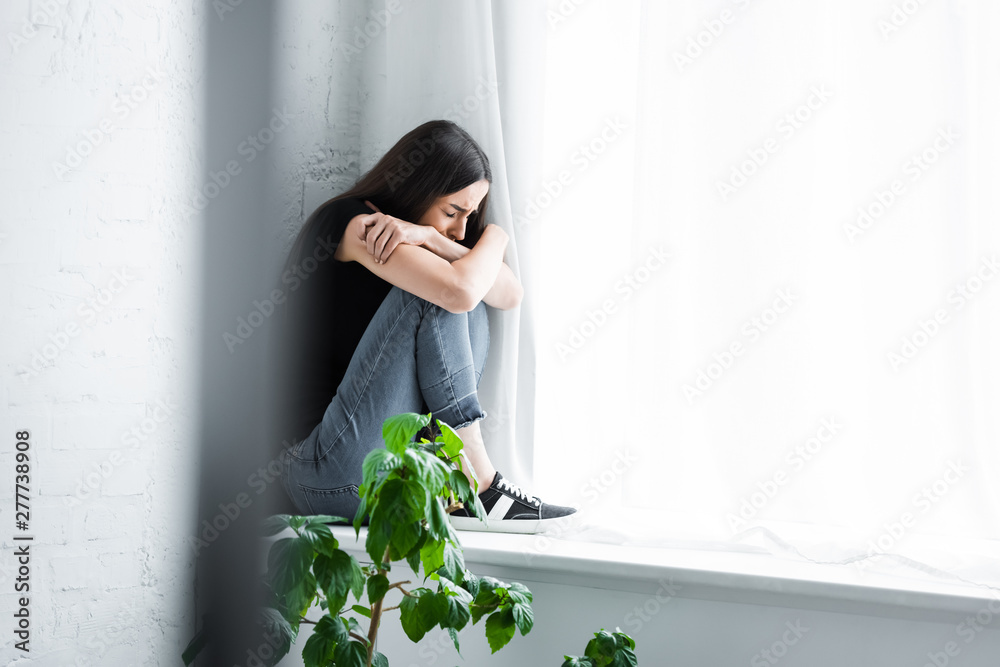 depressed young woman crying while sitting on window sill and hiding ...