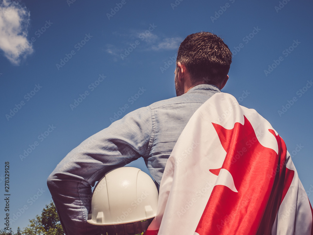 Handsome engineer, holding white hardhat and Canadian Flag in the park ...