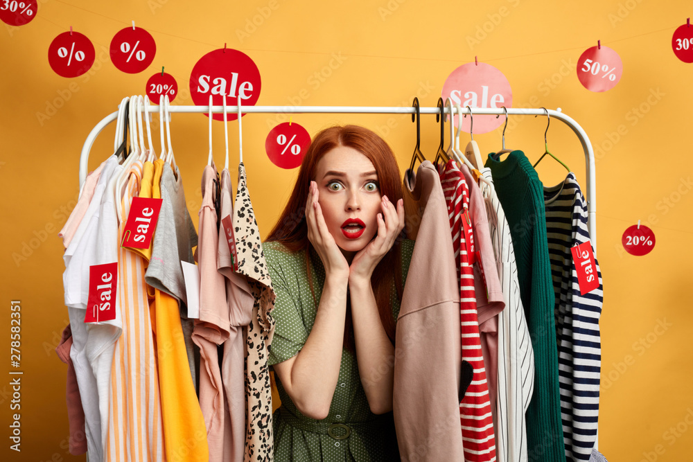surprised girl with palms on her cheek standing among clothes in the ...