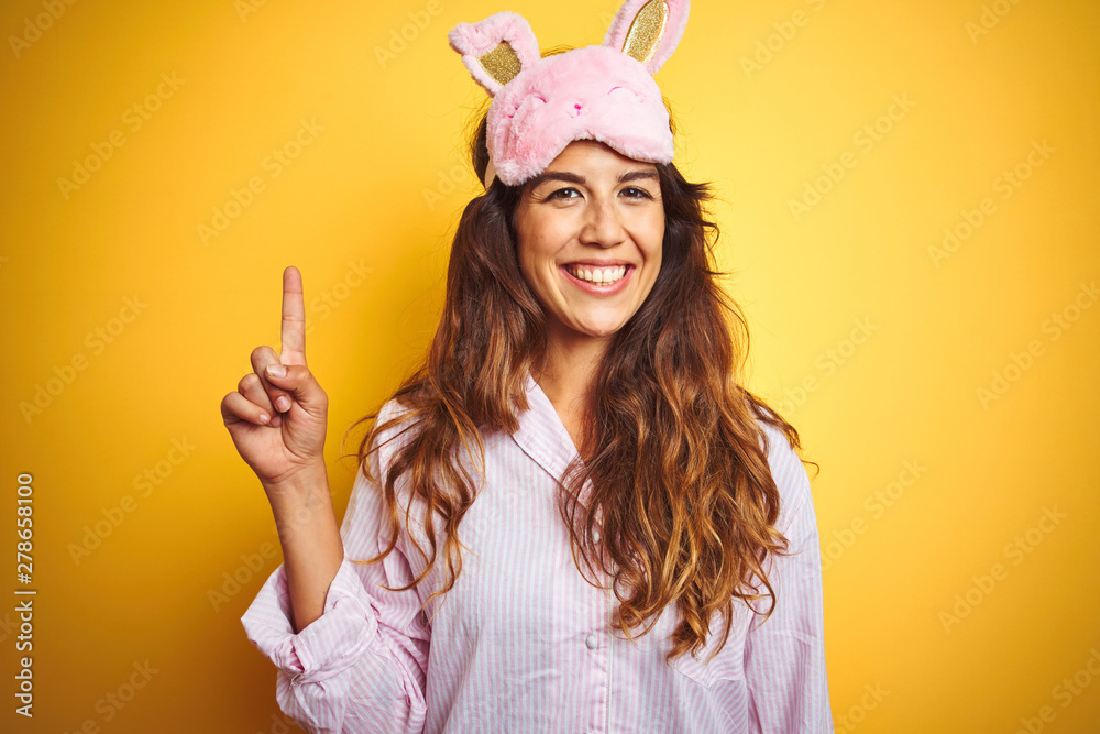 Young woman wearing pajama and sleep mask standing over yellow isolated ...