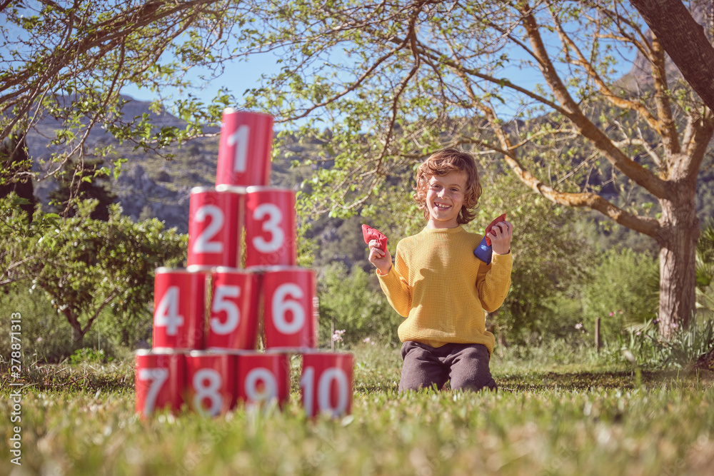 Cheerful kid kneeling and throwing balls in pyramid of red tin cans on ...