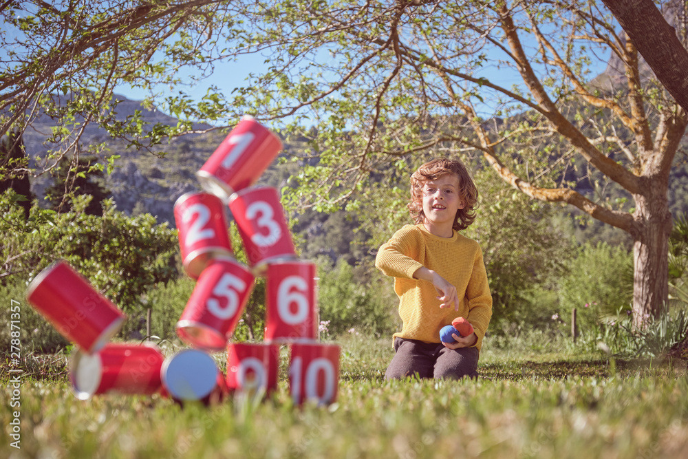 Cheerful kid kneeling and throwing balls in pyramid of red tin cans on ...