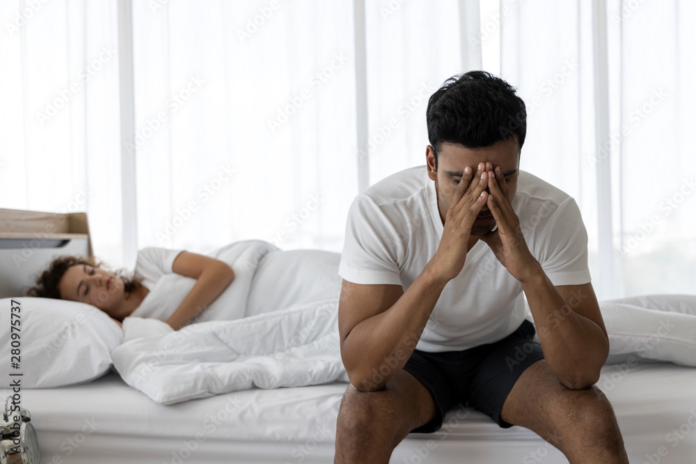 Upset Depressed Young Man Sitting On The Edge Of Bed Against His Wife