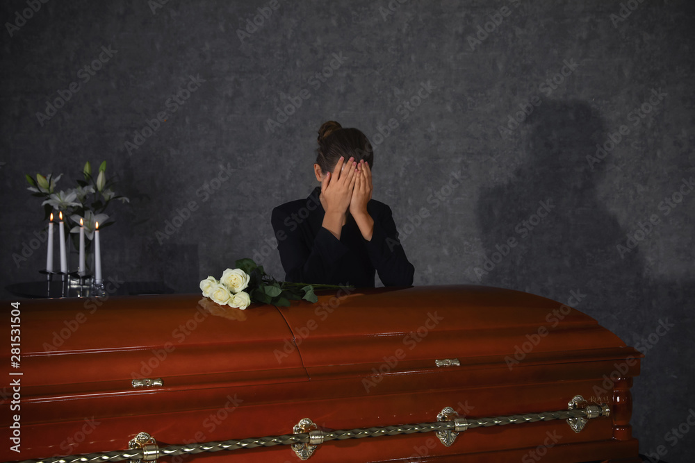 Sad young woman mourning near casket with white roses in funeral home ...