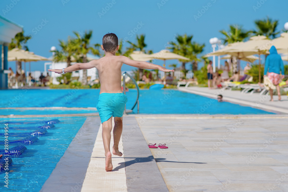 Joyful European boy running along swimming pool at resort in summer ...