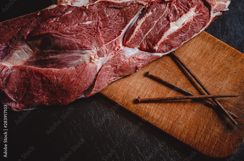 Raw beef shoulder on a dark table. Nearby are three large rusty nails ...