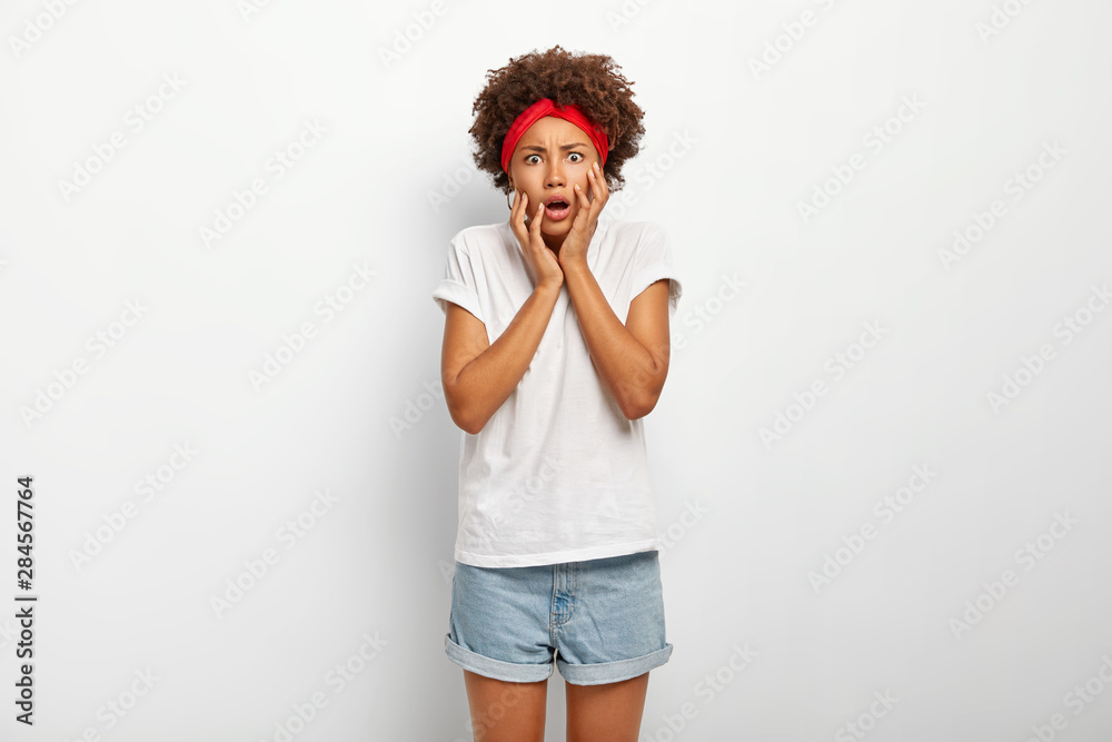 Scared excited curly haired woman looks nervously at camera, touches ...