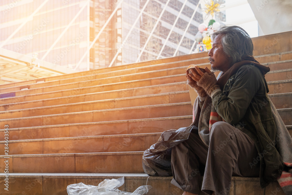 Homeless man is sitting down on staircase in town.He is eat bread and ...