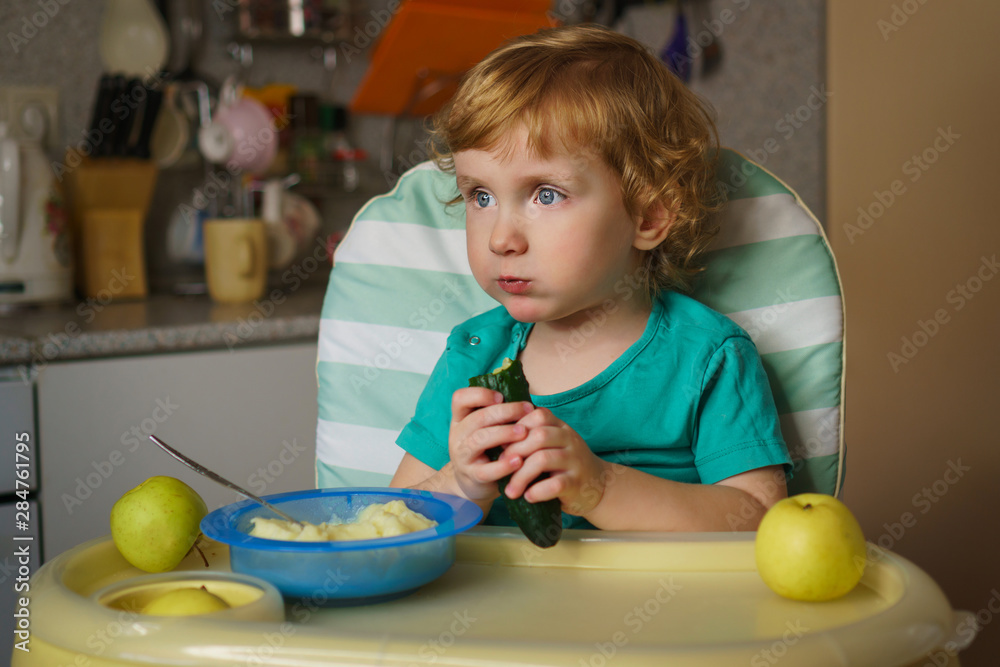 Charming little child eating in the kitchen. He sits in a highchair ...