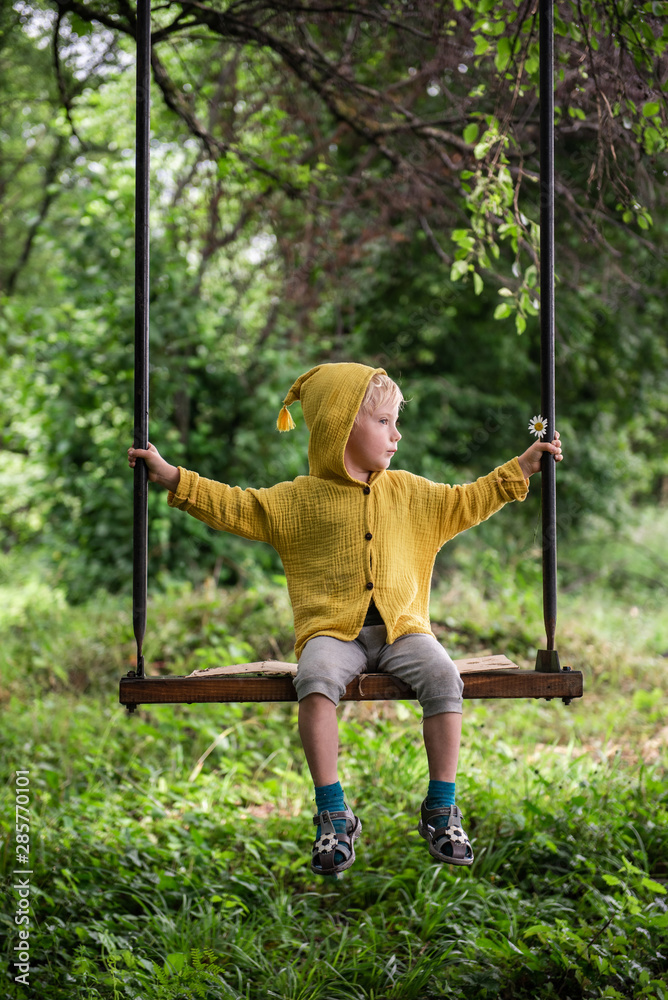 Little boy in a yellow suit swings on a swing on under a tree next to a ...