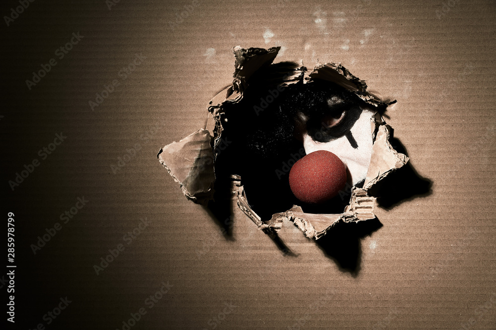 A creepy scary vampire clown with a red nose looks through a torn ...