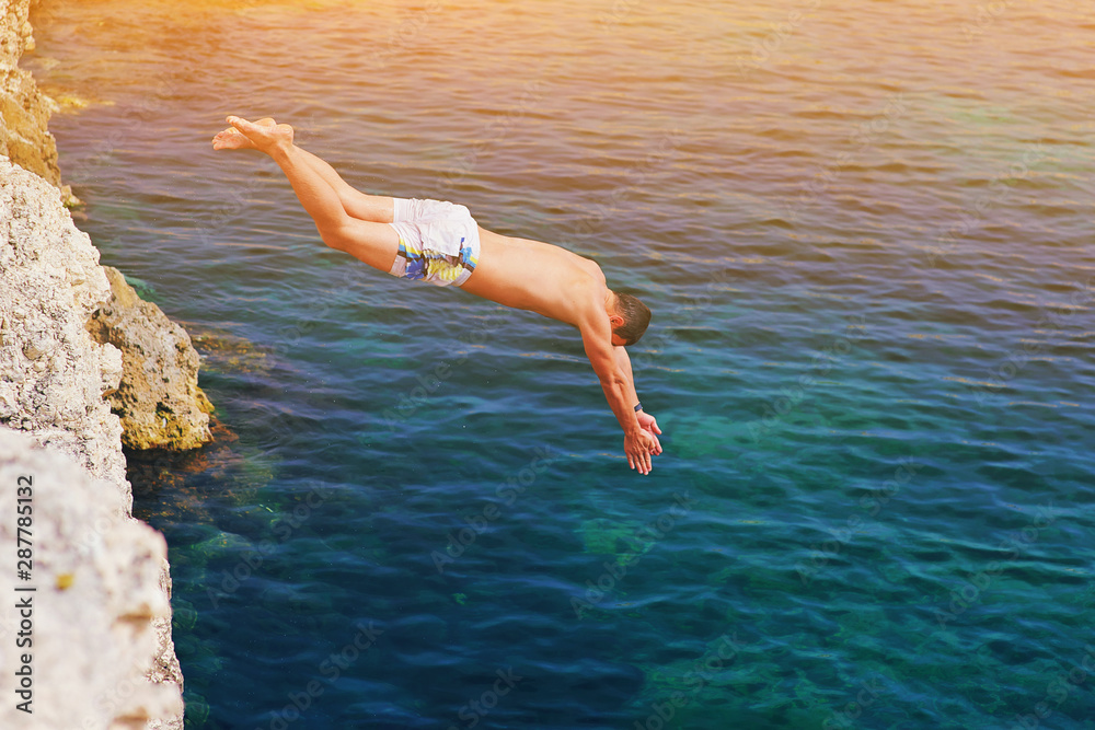Young man jumping off cliff into blue water ocean at sunset. Active ...