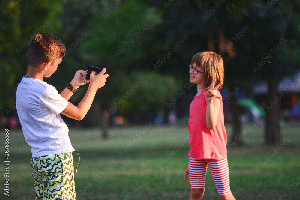 Boy taking photo with cell phone girl while posing in park. A brother ...