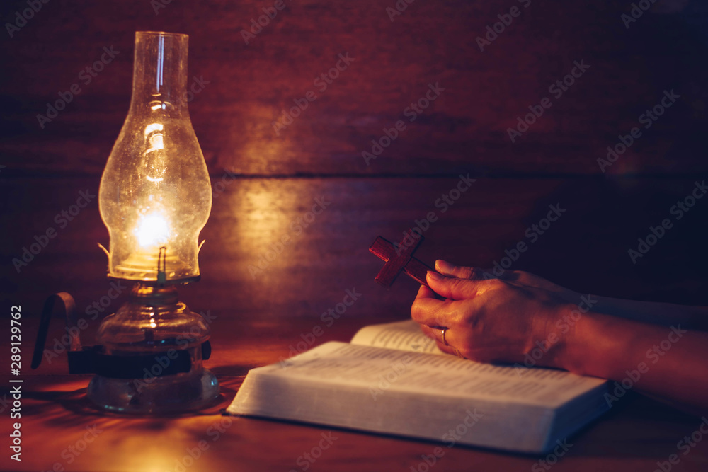 Close up of woman hands hold wooden cross over open bible with oil lamp ...