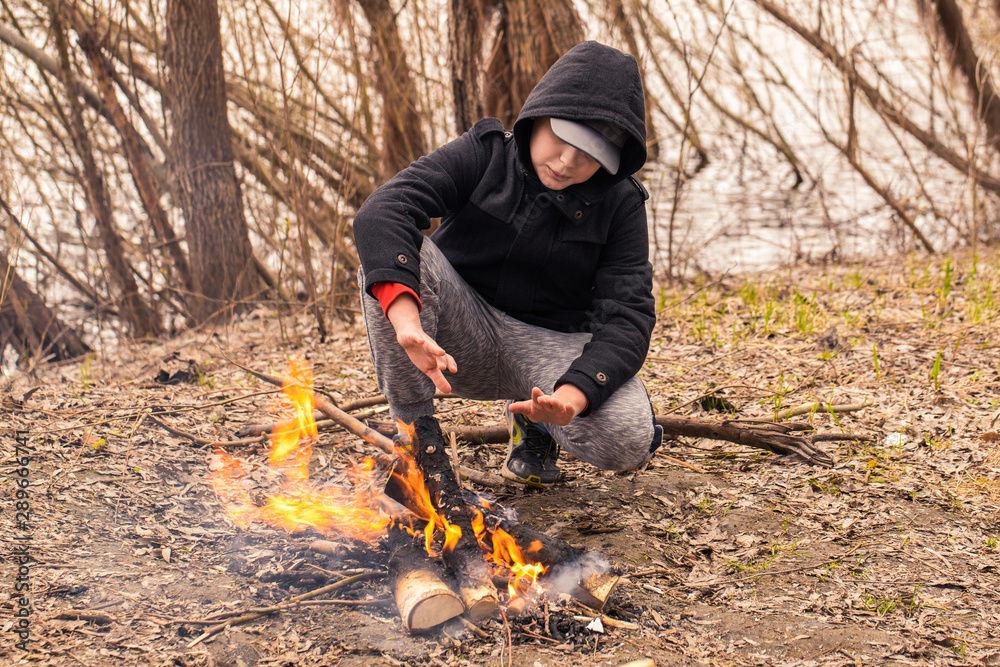 White young kid sitting near burning hot campfire in cold spring forest ...