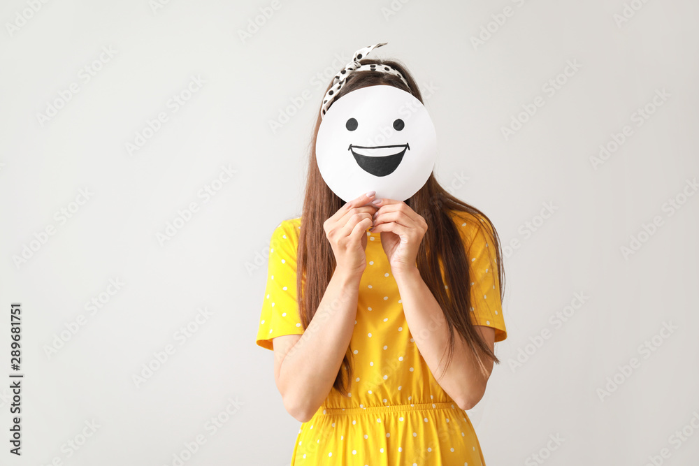 Woman hiding face behind sheet of paper with drawn emoticon on grey ...