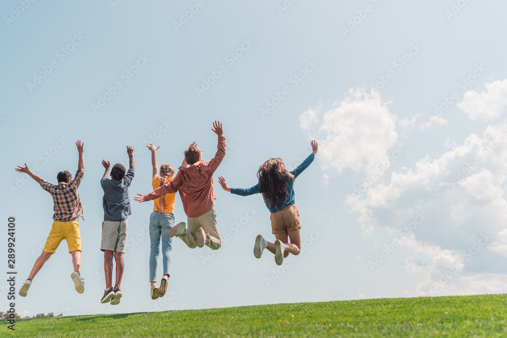 back view of multicultural kids jumping and gesturing against blue sky ...