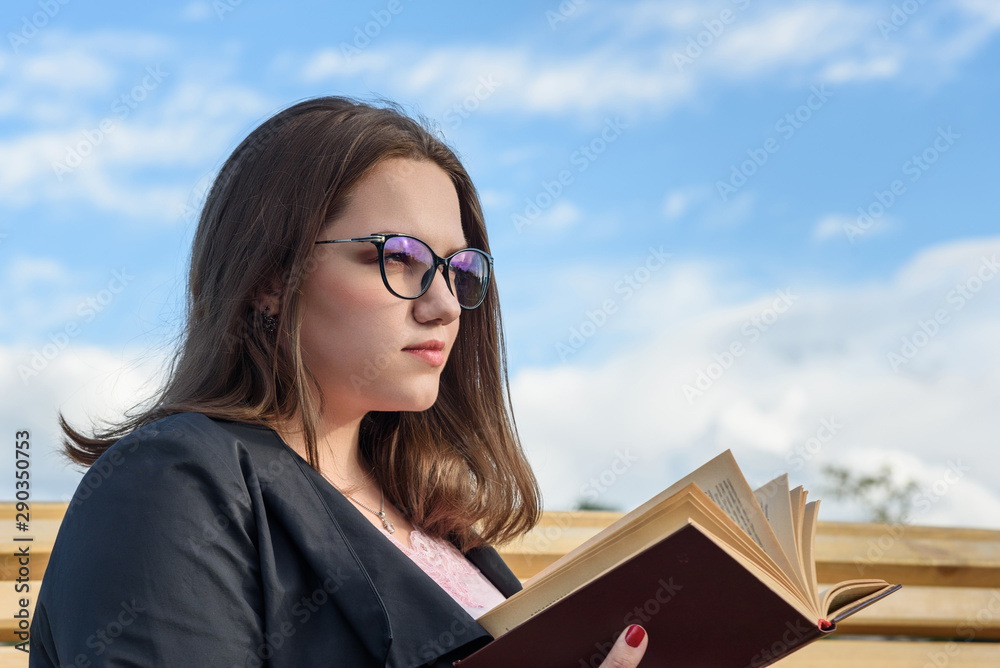 Brunette girl reading book on bench on the street. Girl in black jacket ...