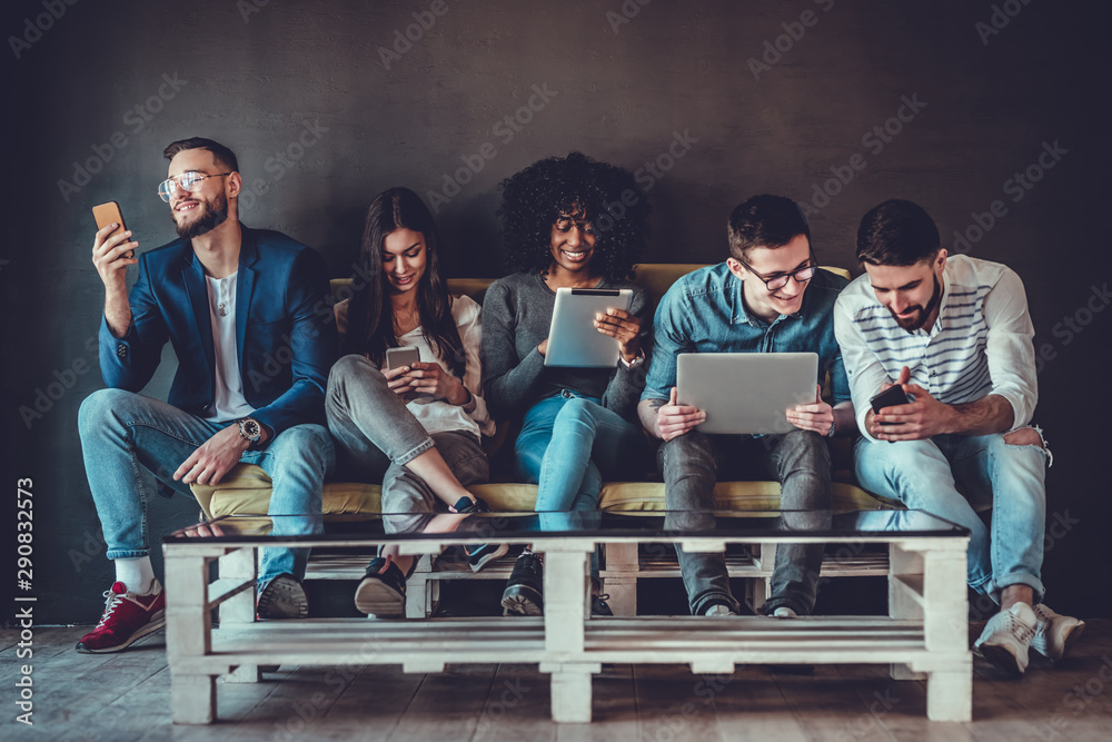 Diverse students using gadgets, sit on sofa in line on grey wall ...
