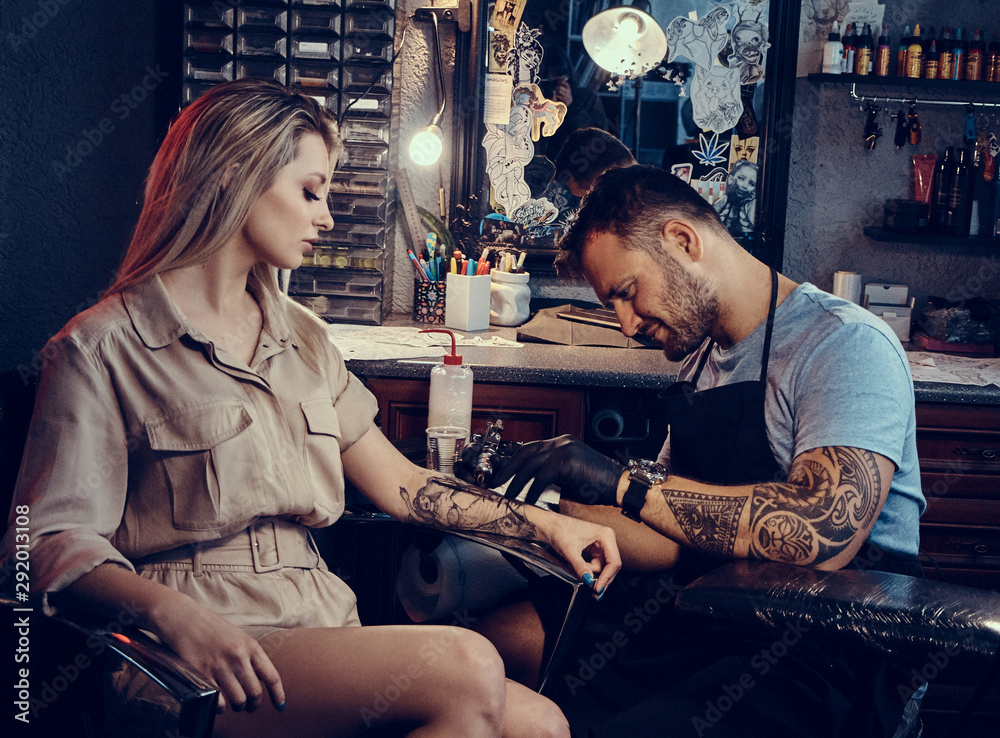 Experienced tattoo master is doing arm tattoo for young woman at his ...