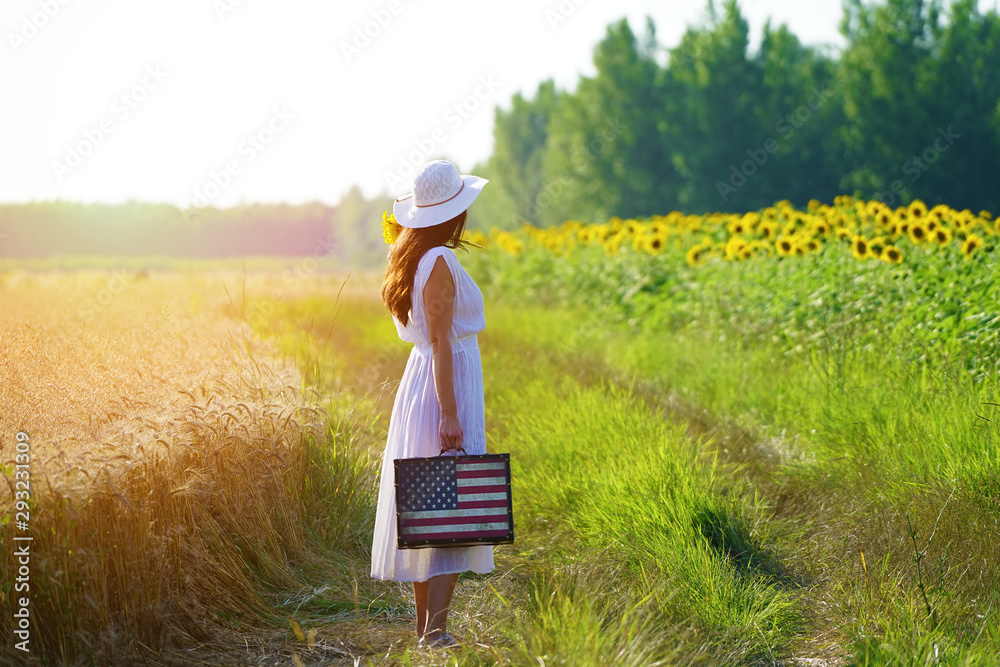 Young woman looking back at golden wheat field, holding sunflowers and ...