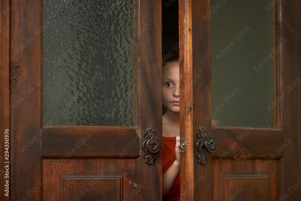 Fotka „A little scared girl is looking through the door slit. A girl is watching someone or ...
