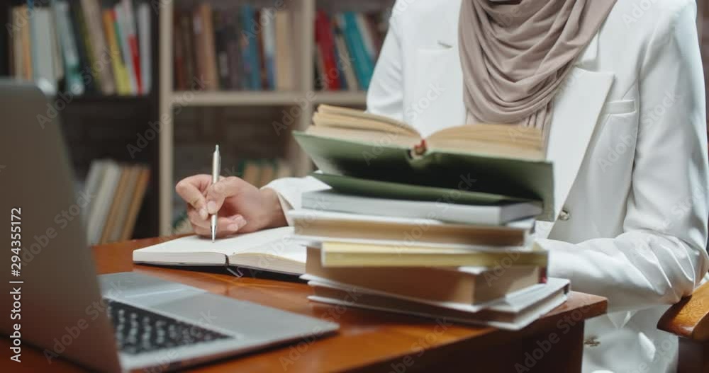 Muslim student is doing research at desk full of books in library. Girl ...