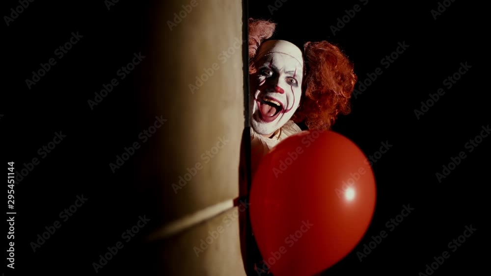 Scary clown with a crazy smile stands around the corner of a dark room ...