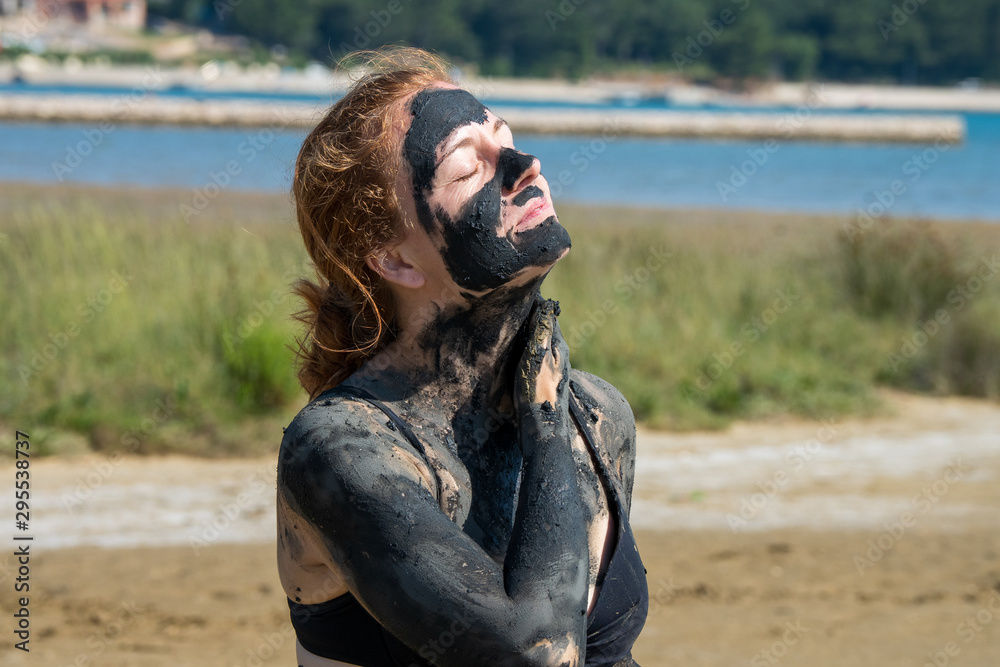 Young woman with ginger hair putting some therapeutic black mud onto ...