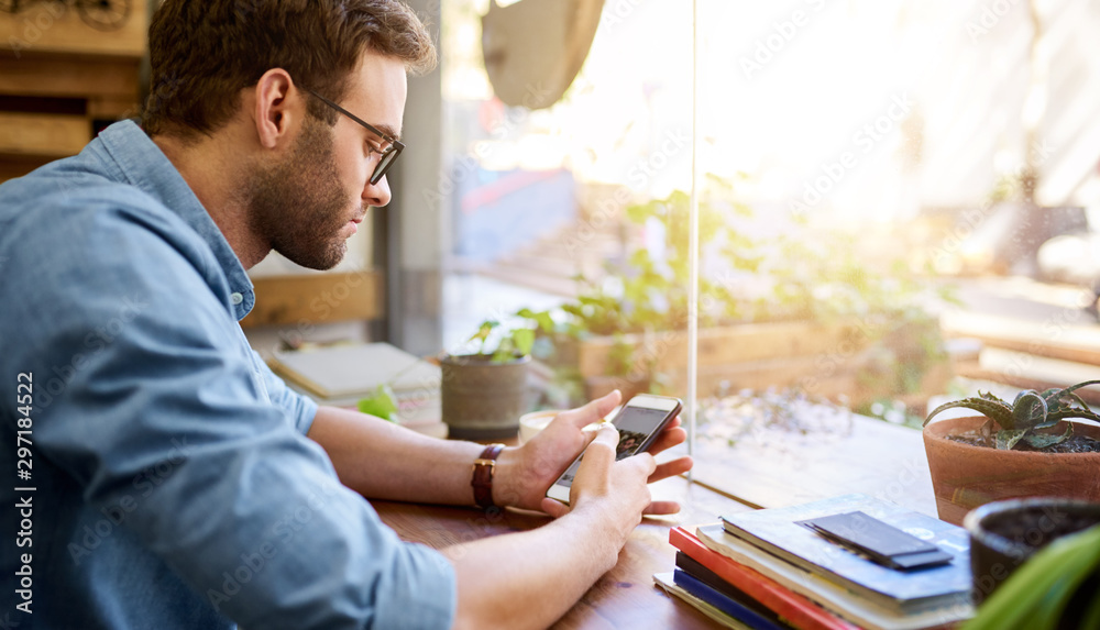 Young man reading a text message on his cellphone while sitting alone ...