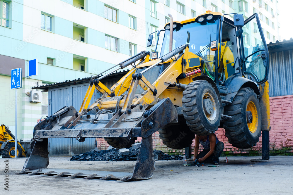 Backhoe loader stands leaning on a bucket. A man repairs a backhoe ...