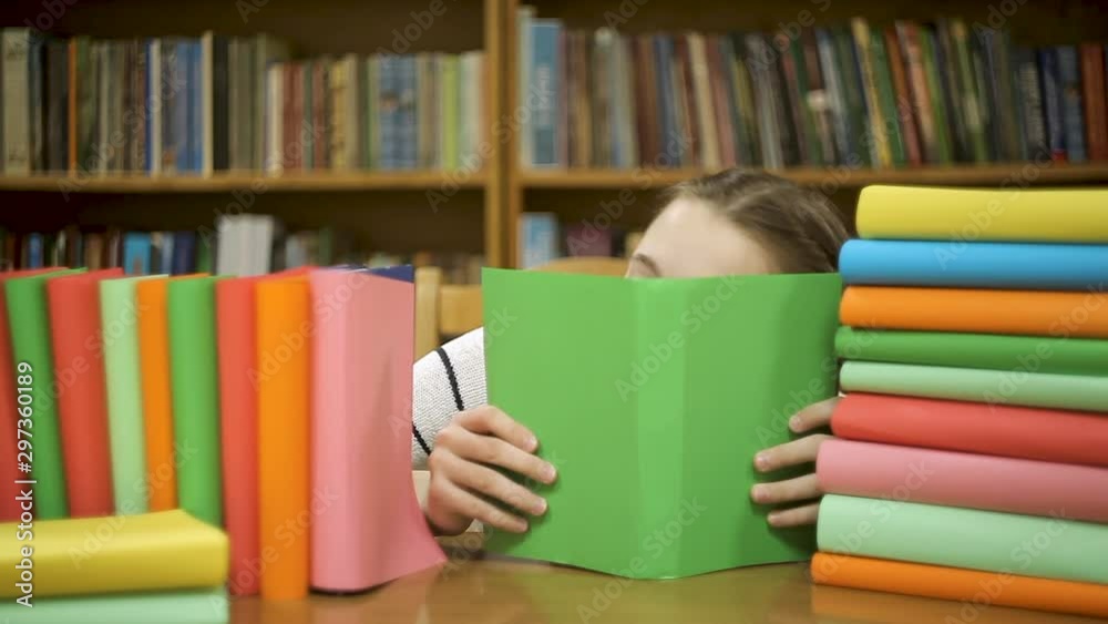 A schoolgirl is hiding behind a book in the library. Children love to ...