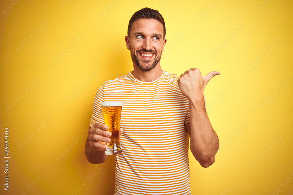 Young handsome man drinking a pint glass of beer over isolated yellow ...