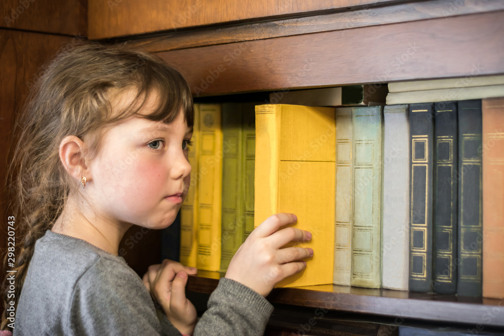Serious little girl is taking a yellow book from the bookshelf. The ...