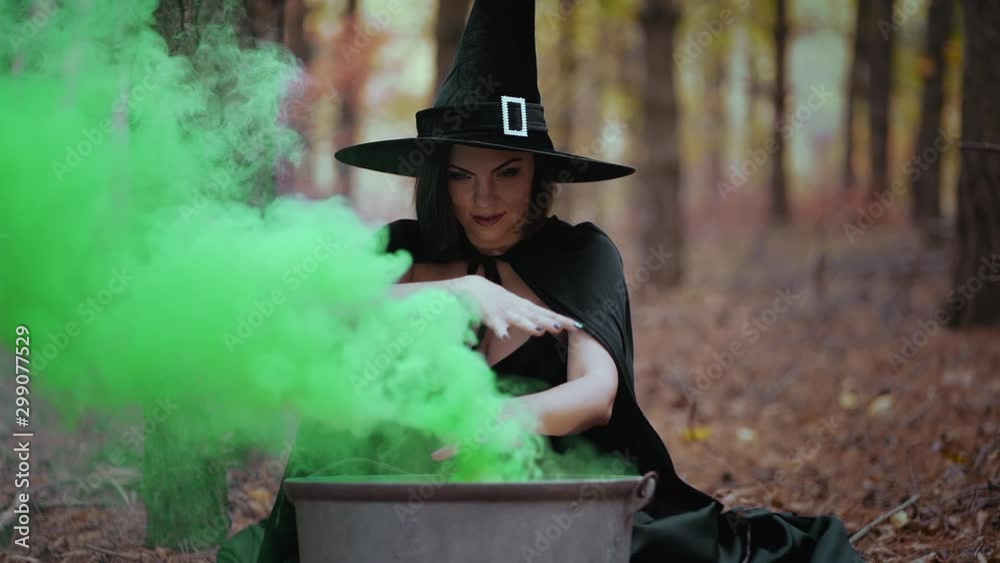 Black witch sits in front of cauldron from which green smoke is falling ...