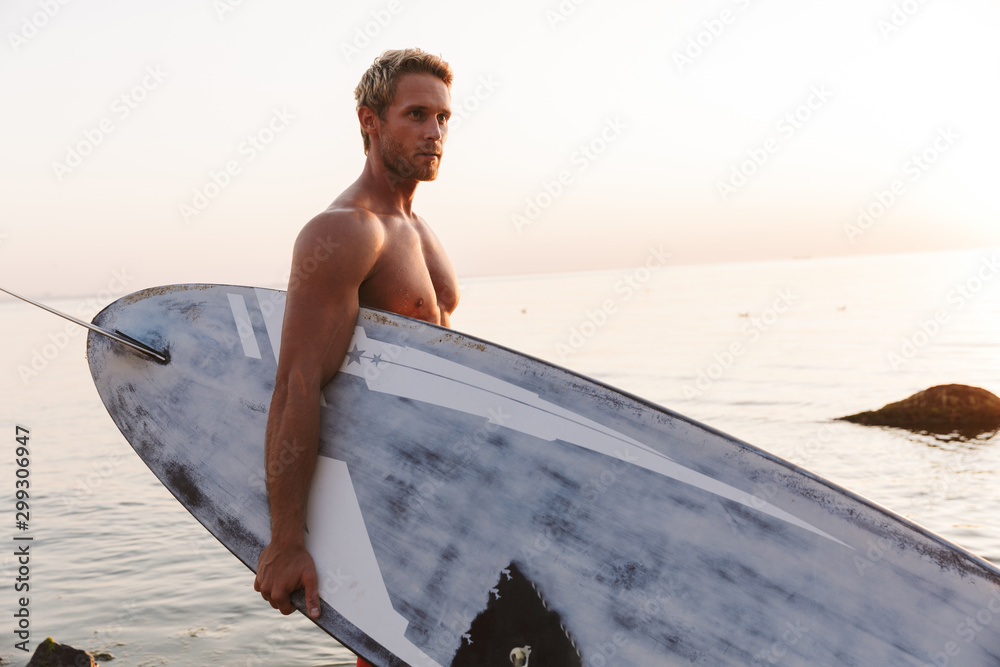 Image of shirtless surfer man holding his surfboard by ocean at sunrise ...
