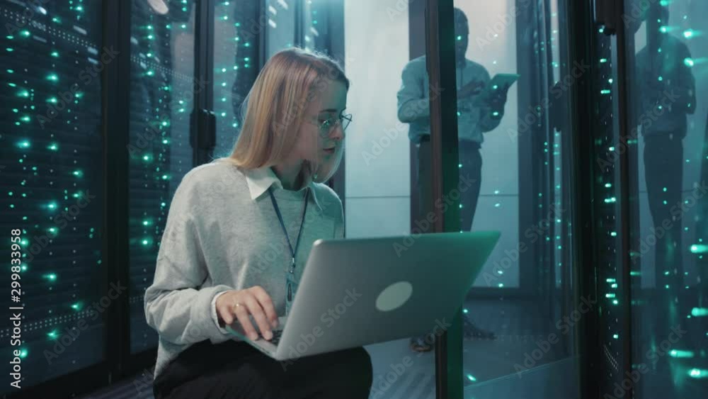 Blonde woman IT administrator working on laptop with open rack server ...