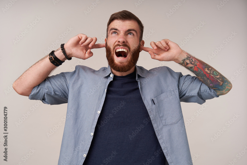 Irritated tattooed man with beard and trendy haircut covering his ears ...