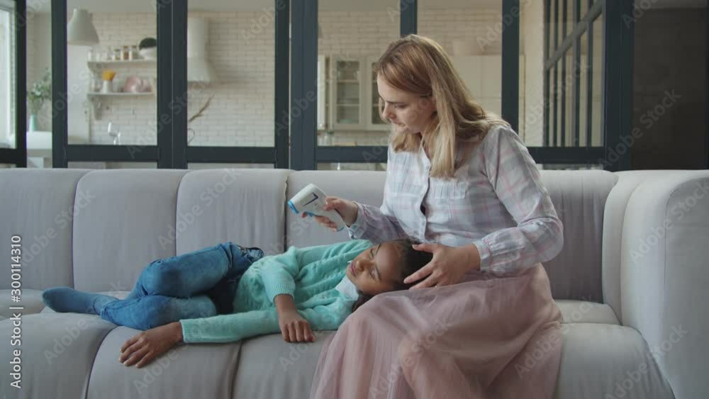 Caring worried mother measuring temperature of sick preadolescent mixed race daughter with ...