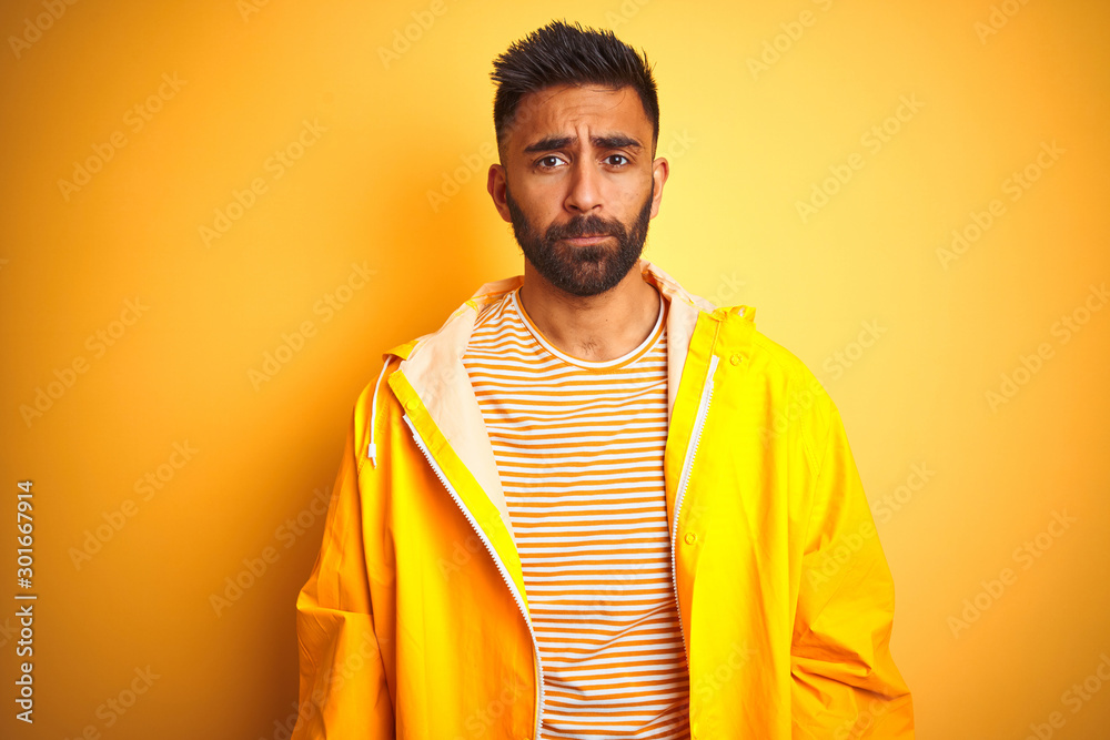 Young indian man wearing raincoat standing over isolated yellow ...