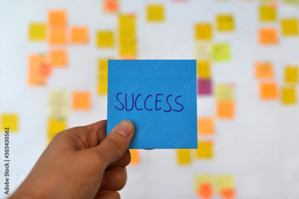 A hand is holding a blue success sticker and there is a Kanban board of ...