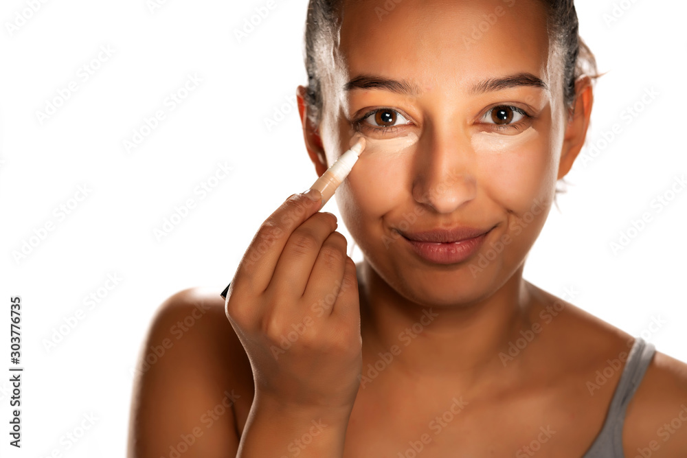 Young dark skinned woman applying concealer on her low eyelids on white ...