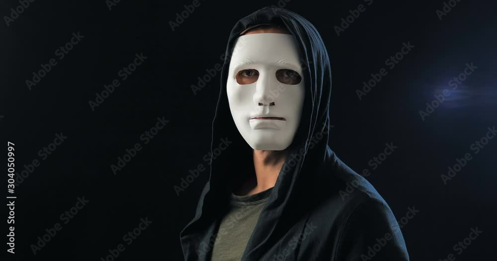 Anonymous man in a white mask covering his face and a black hood turns ...