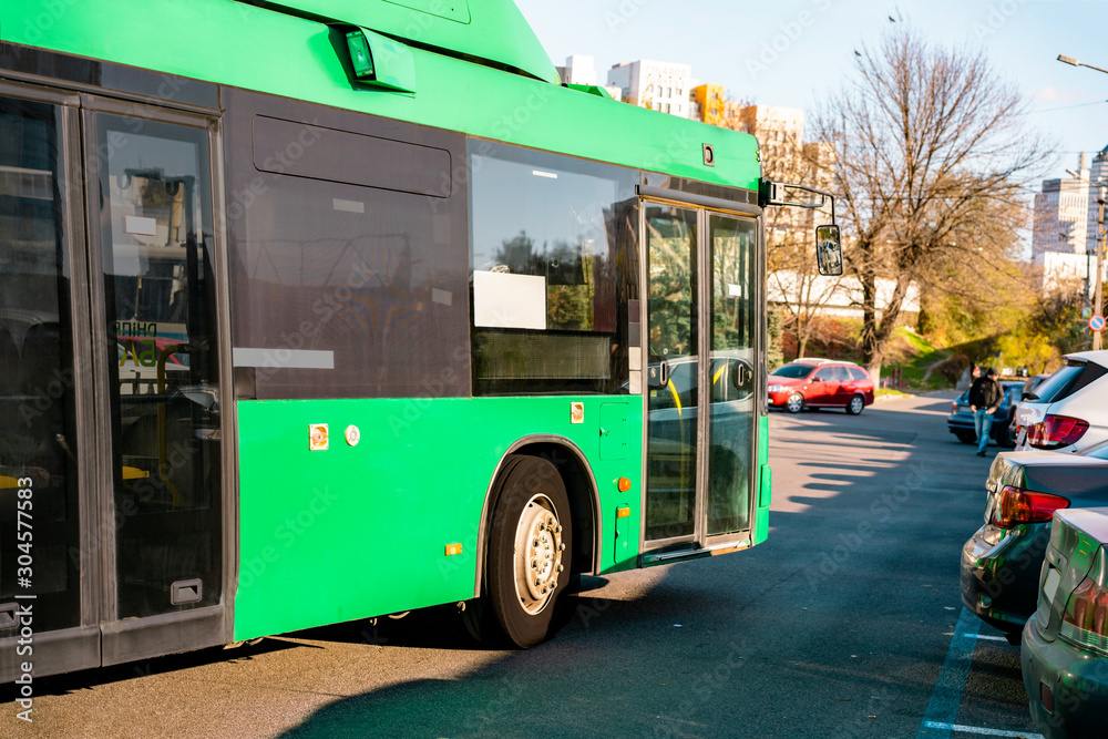 Green electric bus moves along the city's street near the parked cars ...